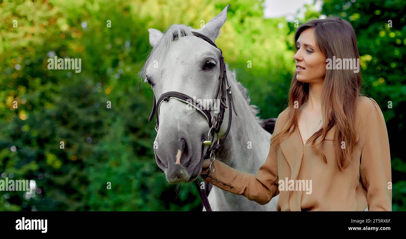 A young woman, a horse rider, guides a saddled grey horse through a ...