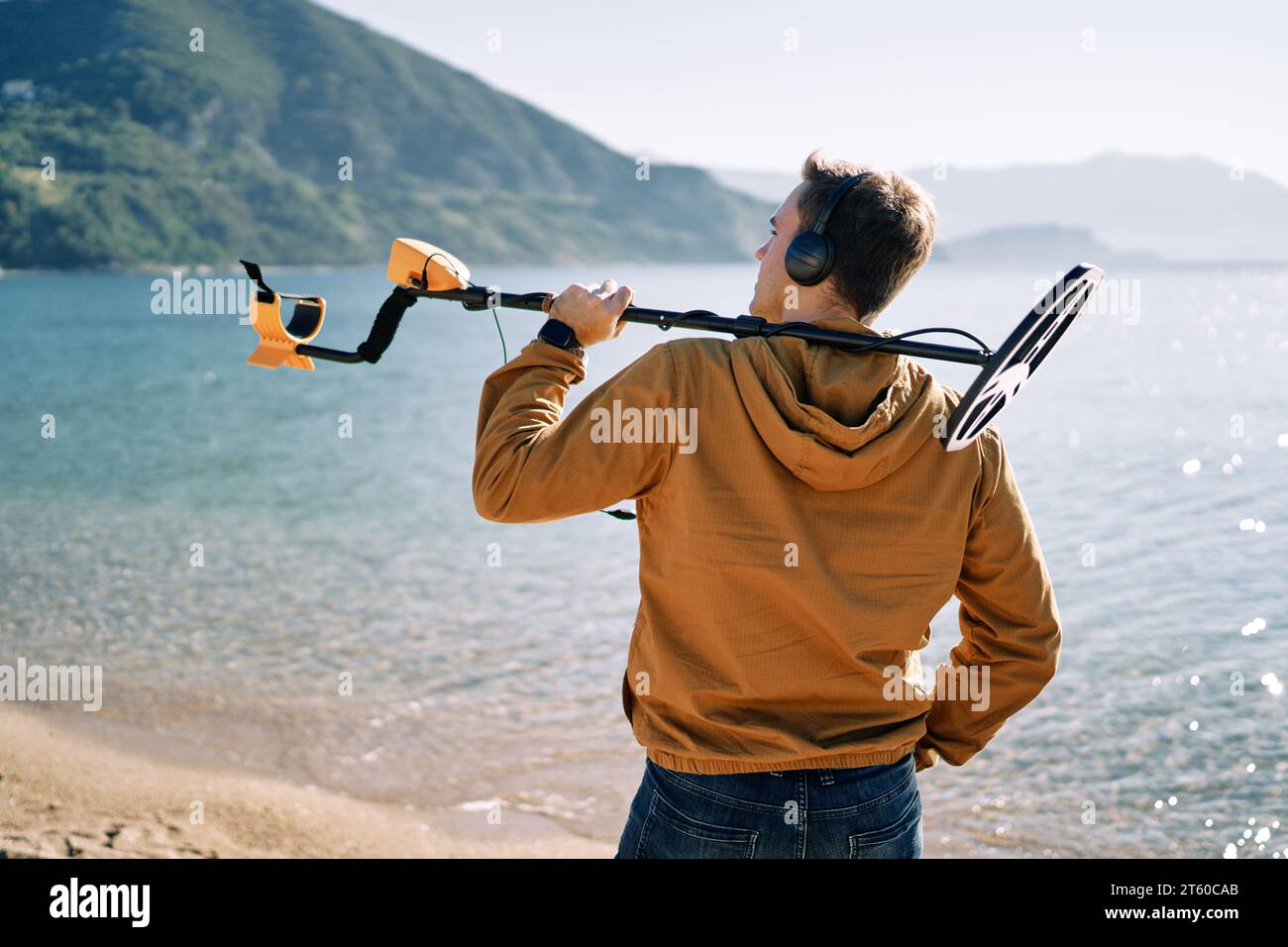 Beach cop stands with a metal detector on his shoulder on the shore and ...