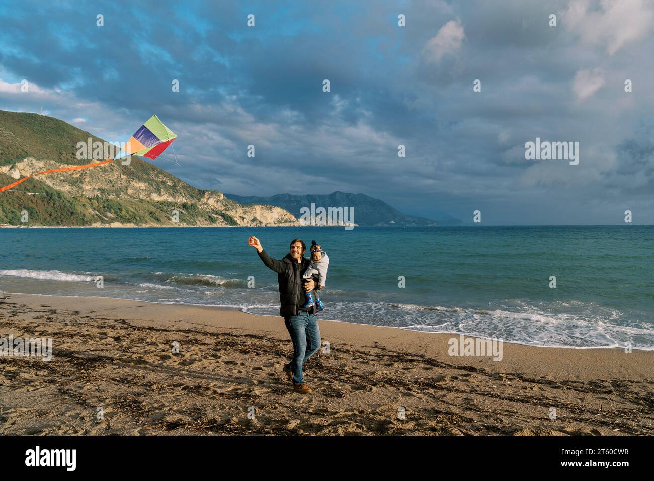 Dad with a little girl in his arms looks at a hovering kite, walking with it along the seashore ...
