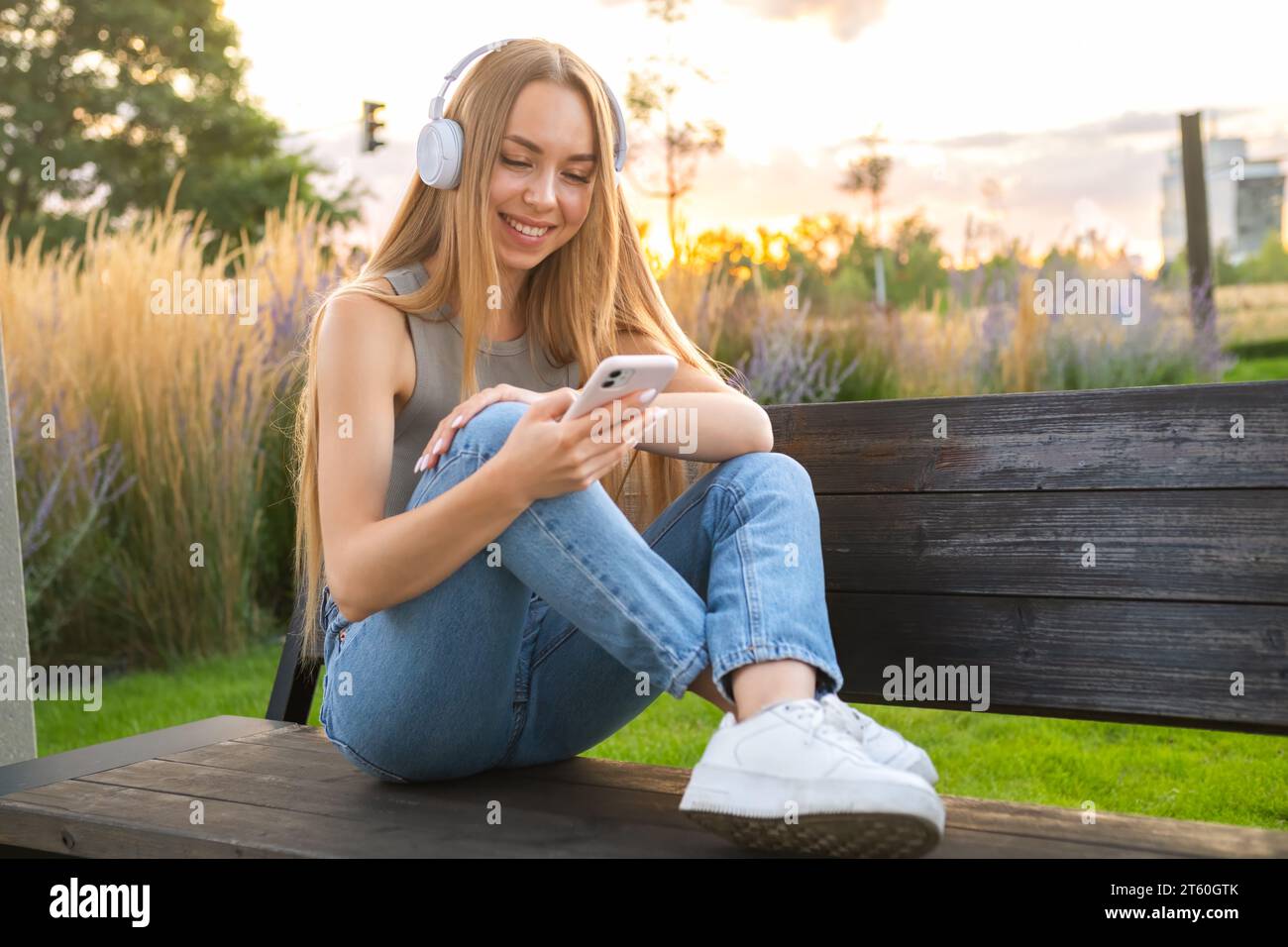 Joyful lady in headphones relaxes hugging legs on park bench in summer ...