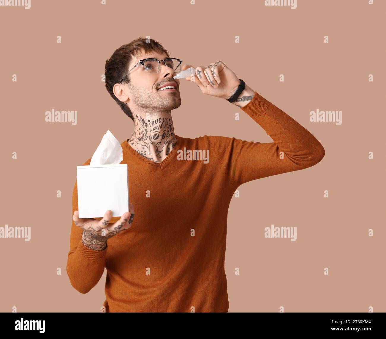 Allergic young man with tissues using nasal drops on brown background ...