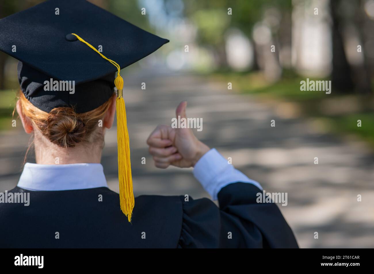 Back view of caucasian woman in graduate gown showing thumb up outdoors ...