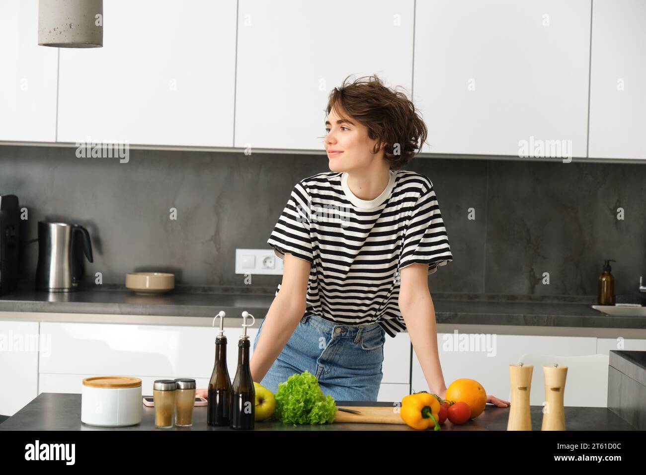 Portrait of young modern woman in the kitchen, leaning on counter with ...