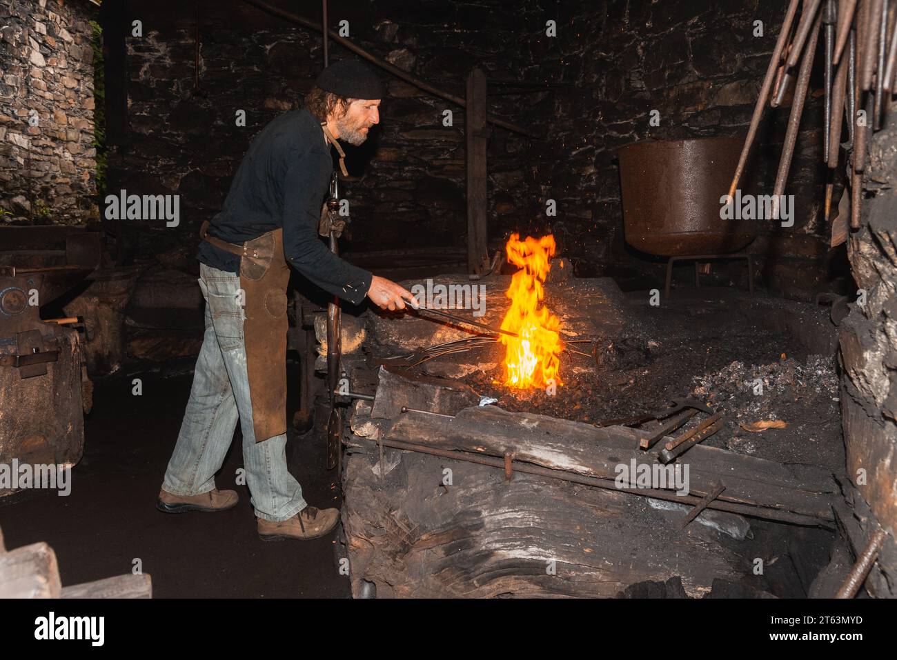 Side view of focused blacksmith, bent over the roaring forge shapes an ...