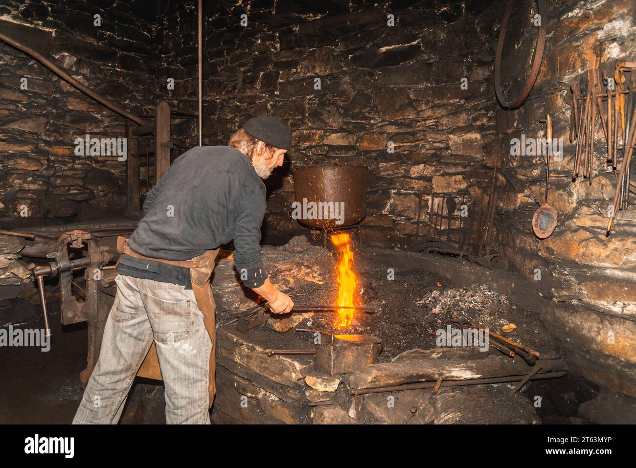 Side view of focused blacksmith, bent over the roaring forge shapes an ...