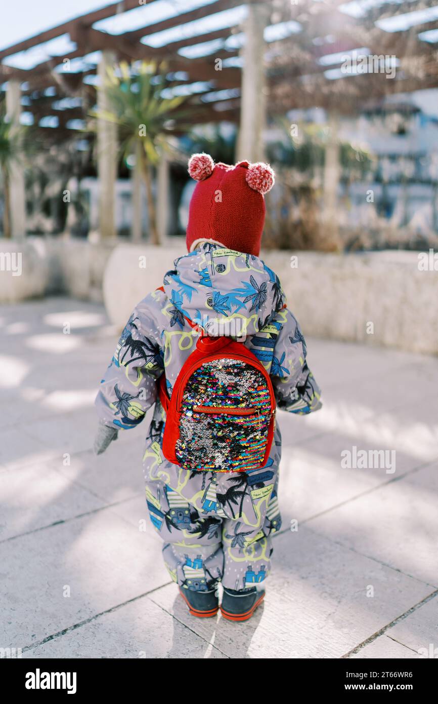 Small child in overalls with a backpack stands on the tiles in the ...