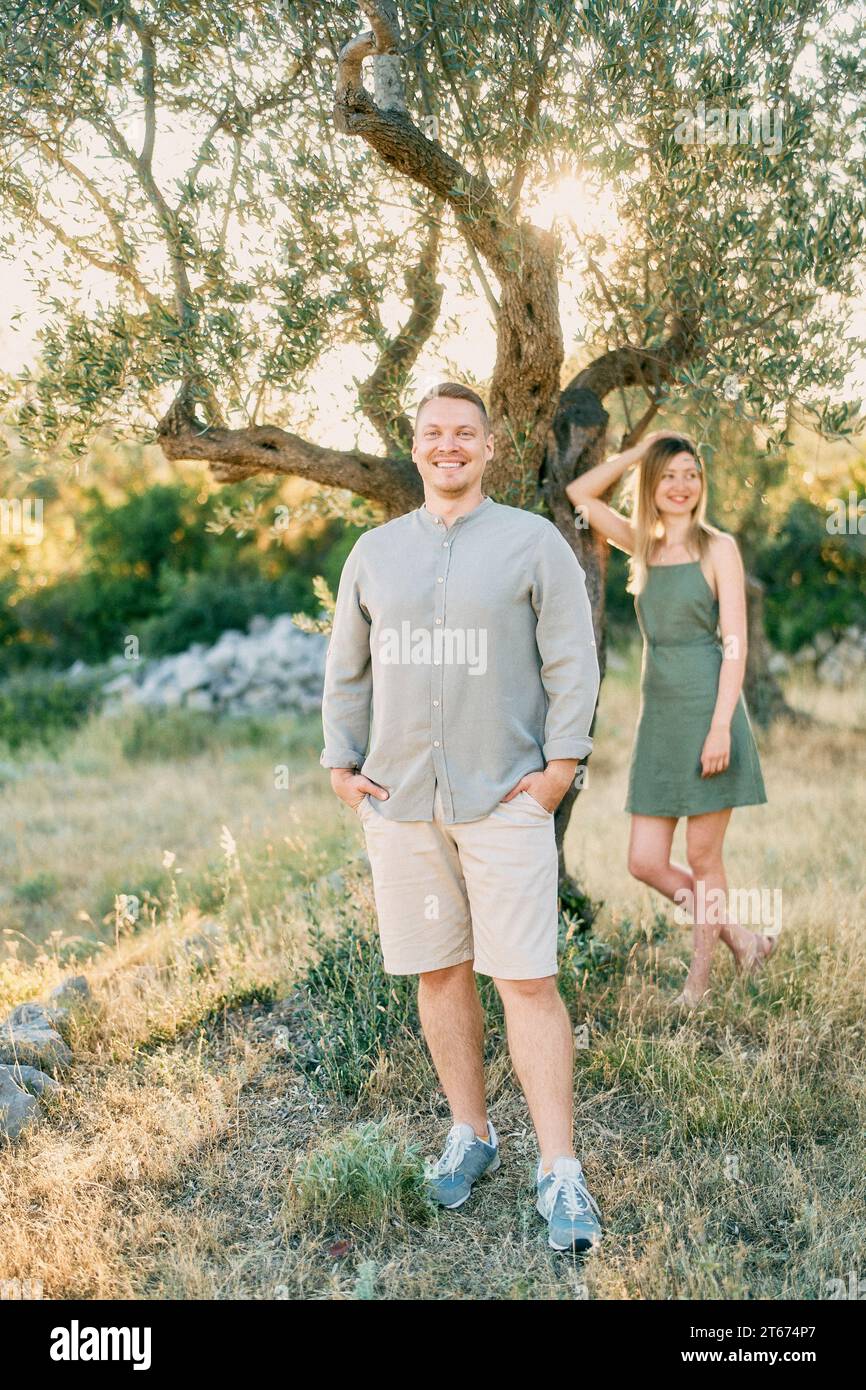 Girl stands leaning her elbow on a tree behind smiling guy with his ...