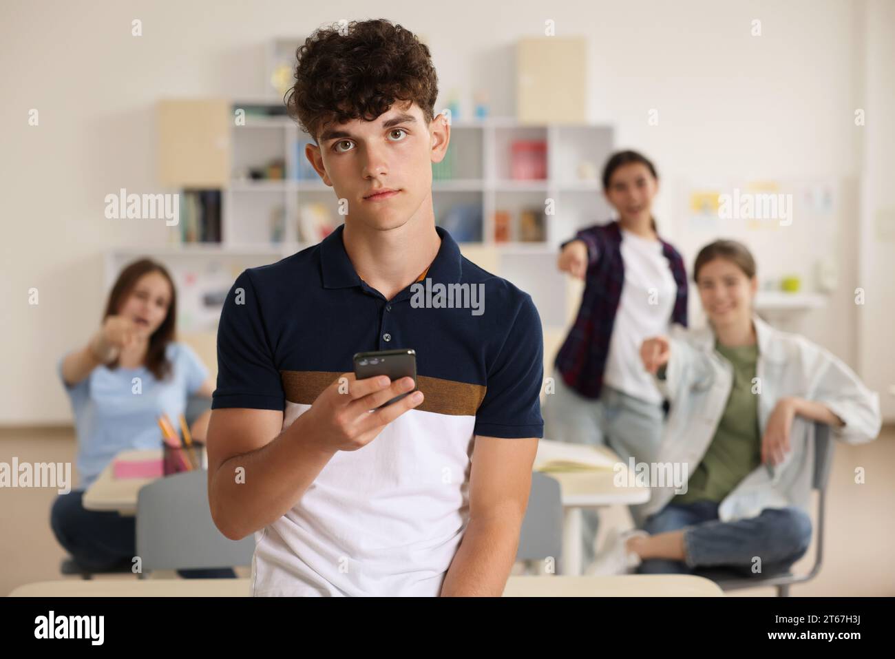 Teen problems. Lonely boy with smartphone standing separately from other students in classroom ...