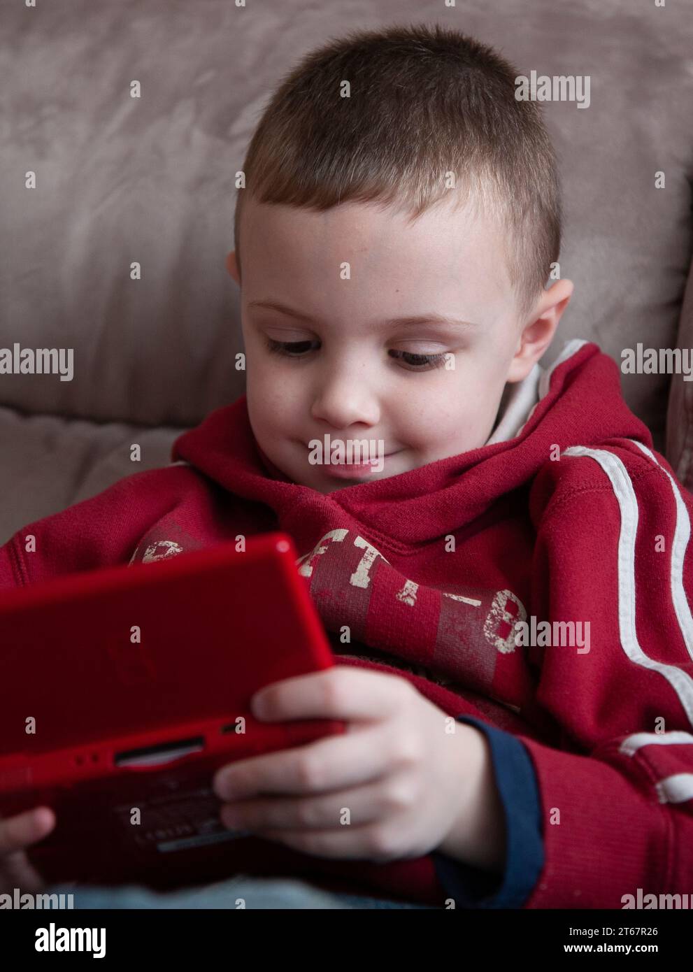 Young boy wearing a bright red hoody top sitting on a beige sofa and ...