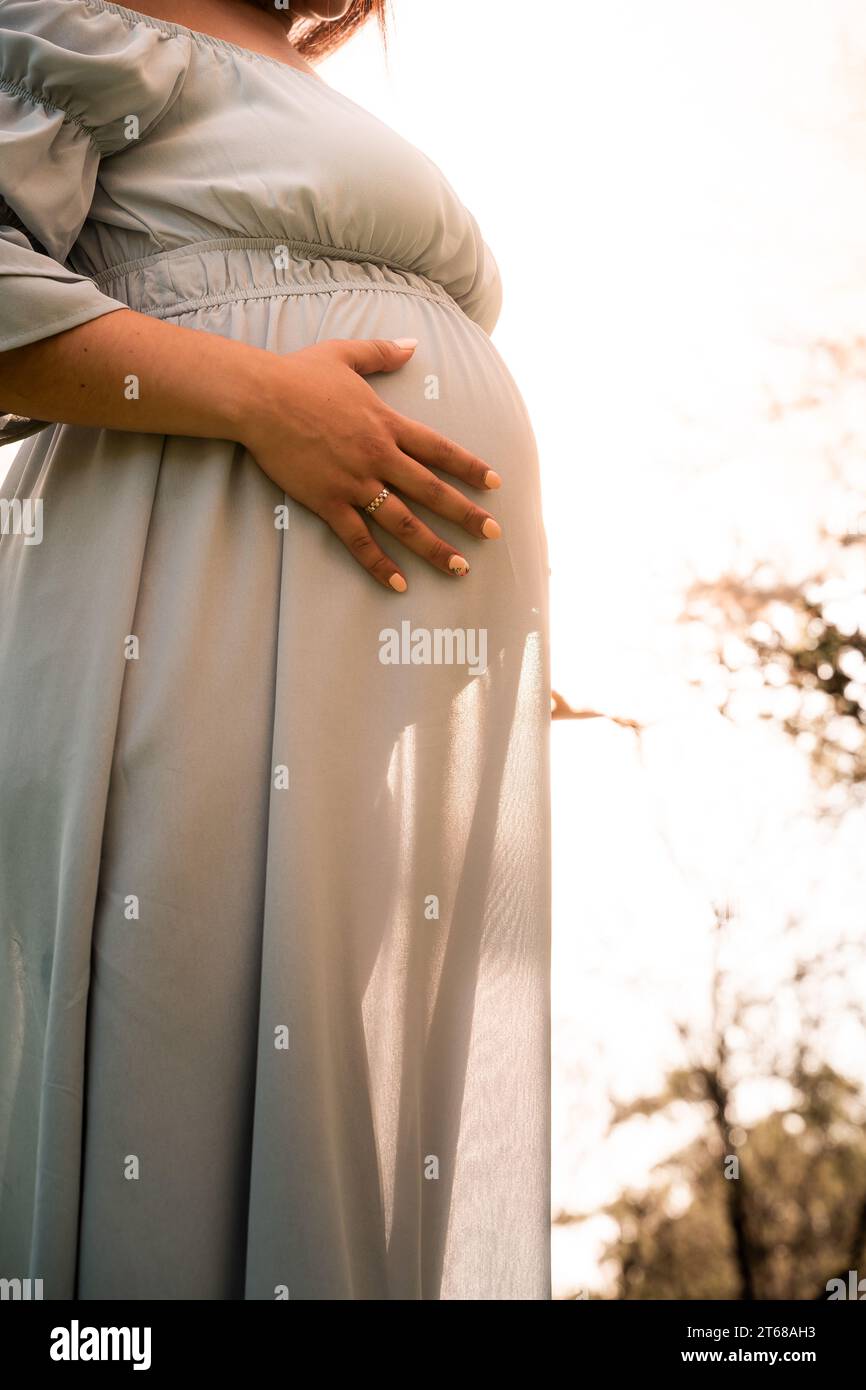 A pregnant Latina woman stands in a park, hands gently cradling her ...