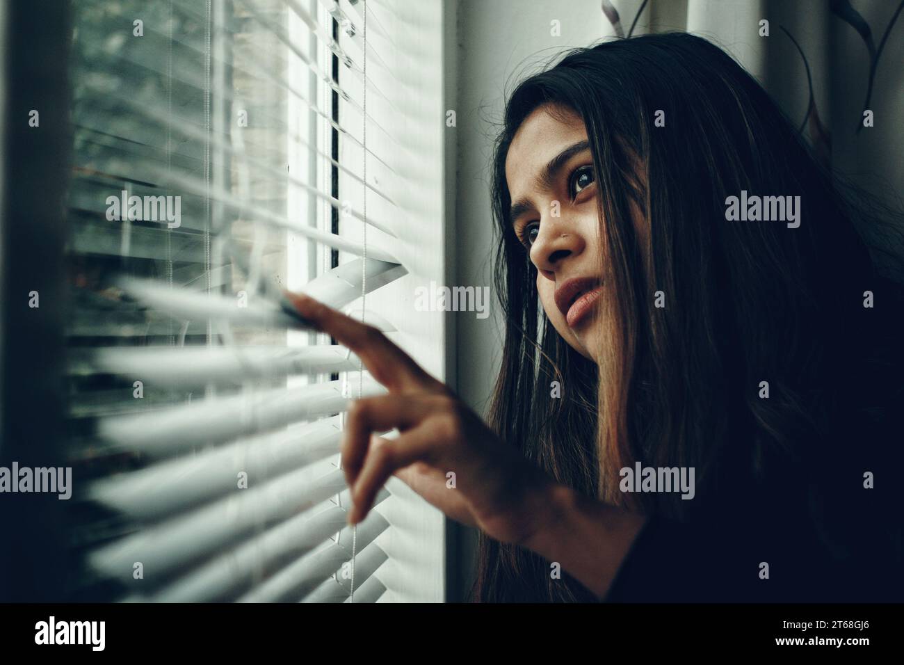 A young female standing at a window, holding white Venetian blinds in ...