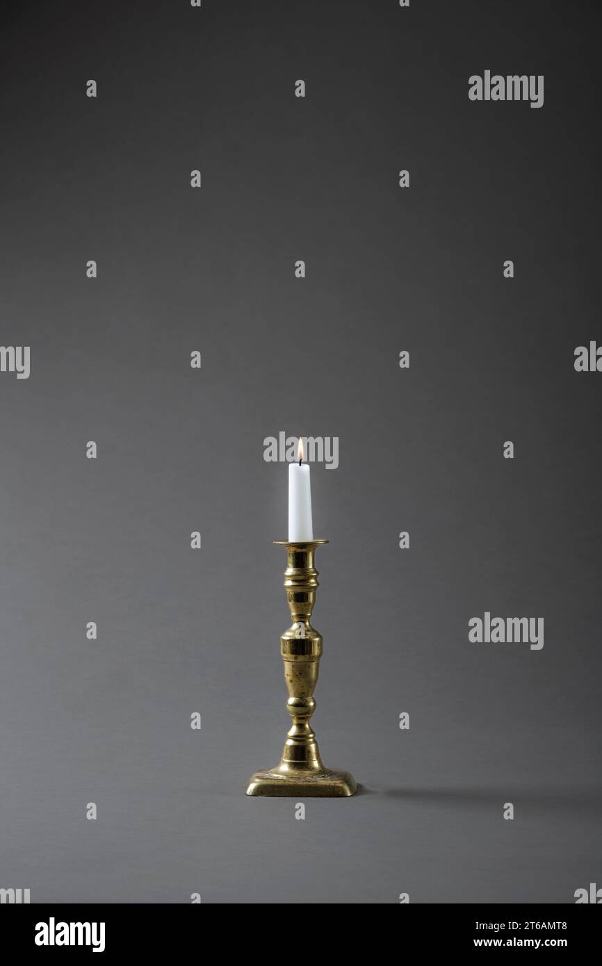 An old brass candlestick with a lit candle, on a plain grey studio ...