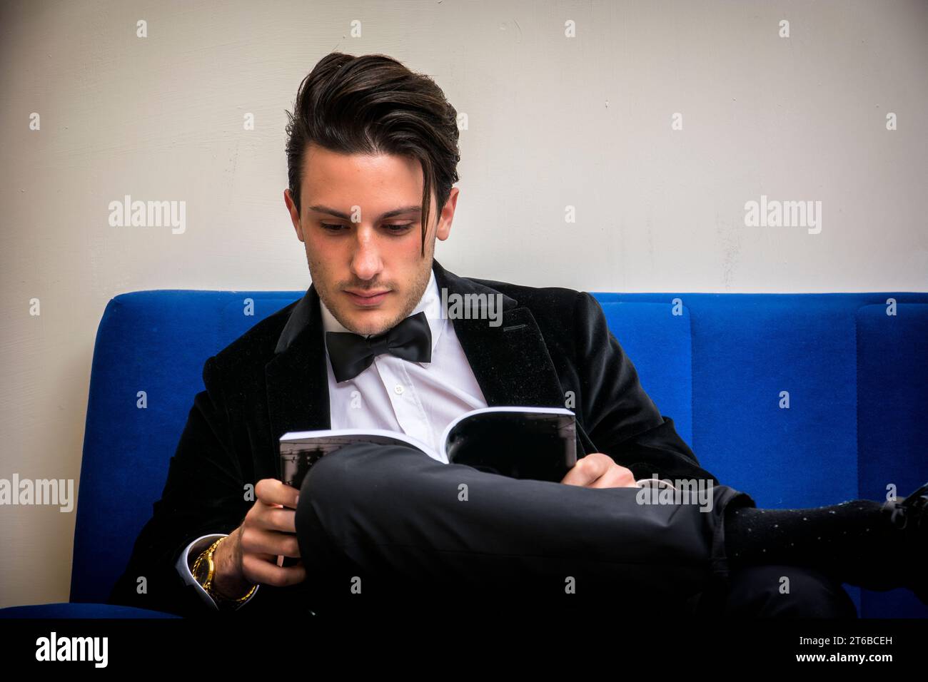 A man in a tuxedo sitting on a blue couch reading a book Stock Photo ...