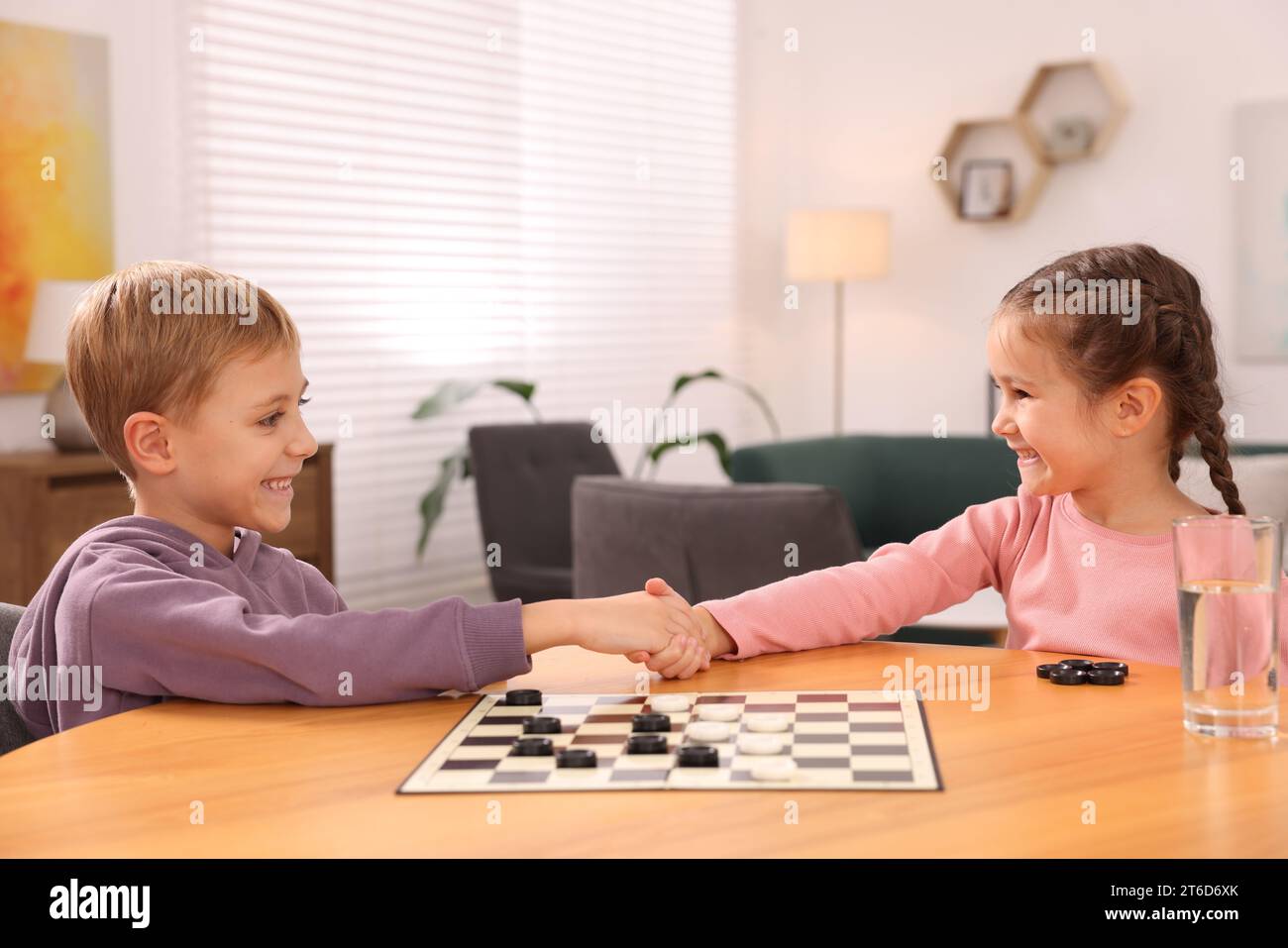 Cute children shaking hands after playing checkers at wooden table in ...