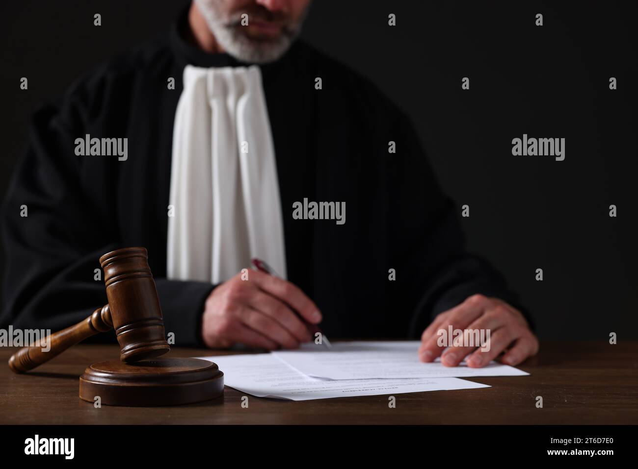 Judge with gavel writing in papers at wooden table against black ...