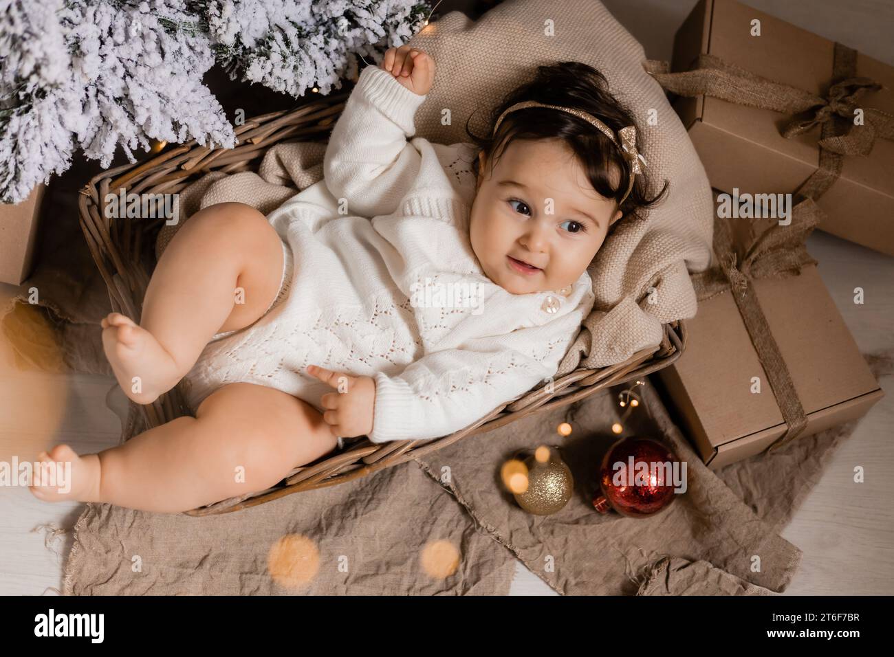 cute baby in white bodysuit lies in a cradle next to the Christmas tree ...