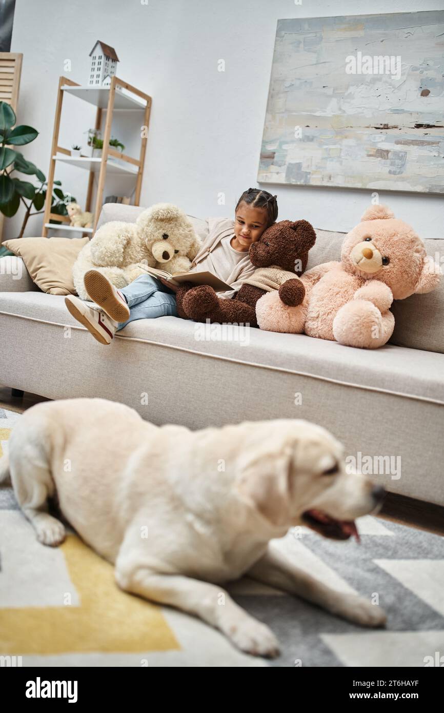 adorable girl sitting on sofa with soft teddy bears and reading book ...