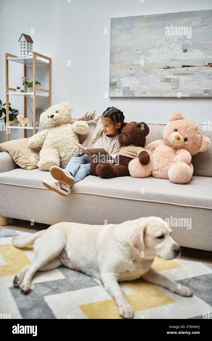 adorable girl sitting on sofa with soft teddy bears and reading book ...