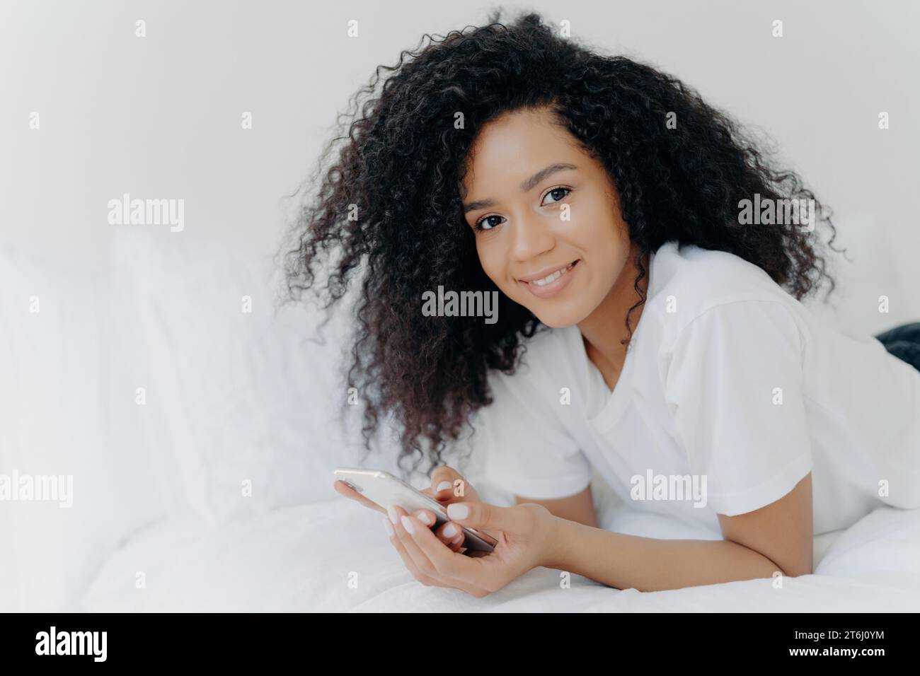 Relaxed young woman with curly hair using smartphone in bed, exuding a ...