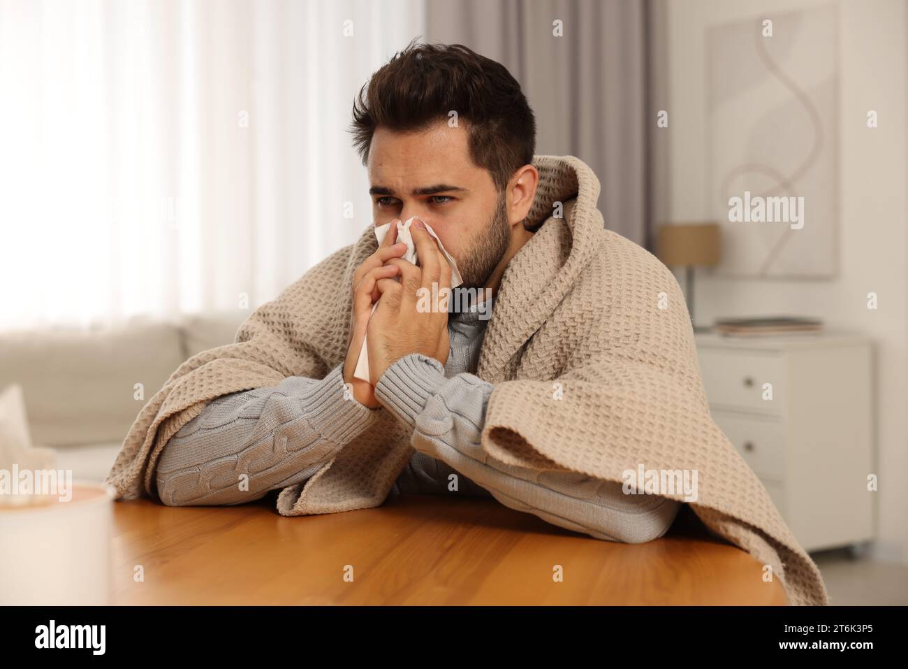 Sick man wrapped in blanket with tissue blowing nose at wooden table ...
