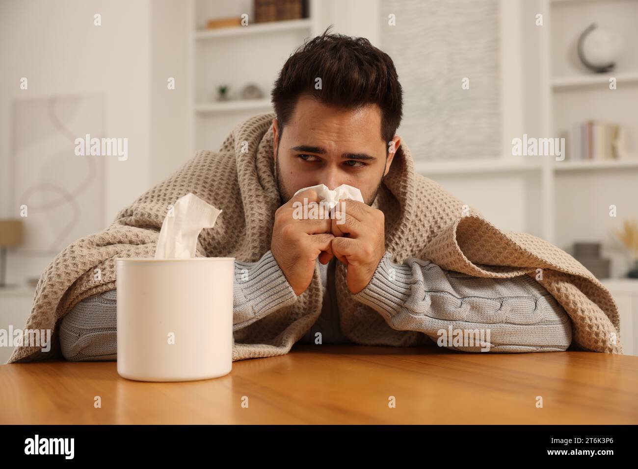 Sick man wrapped in blanket with tissue blowing nose at wooden table ...