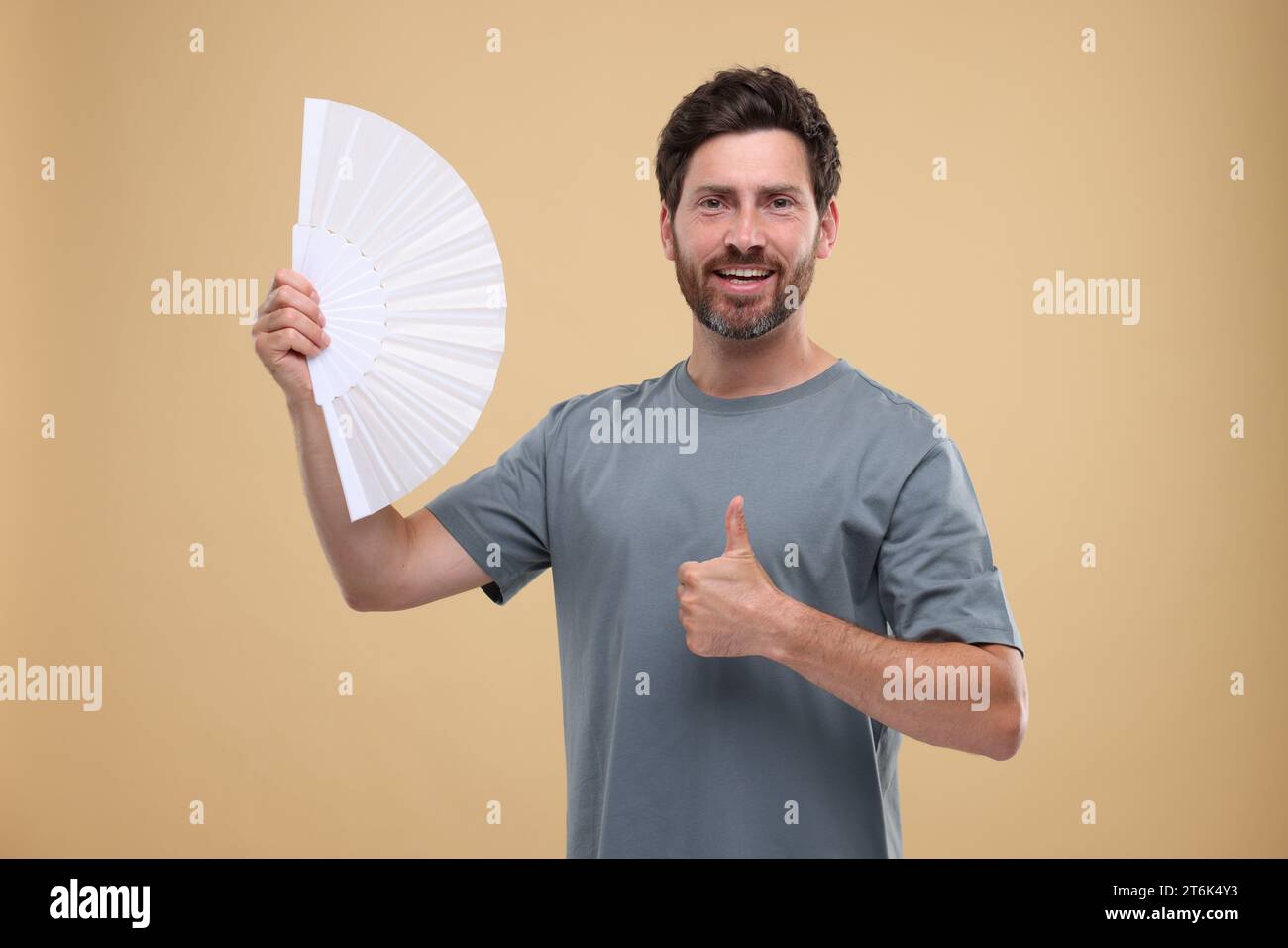 Happy man holding hand fan and showing thumb up on beige background ...