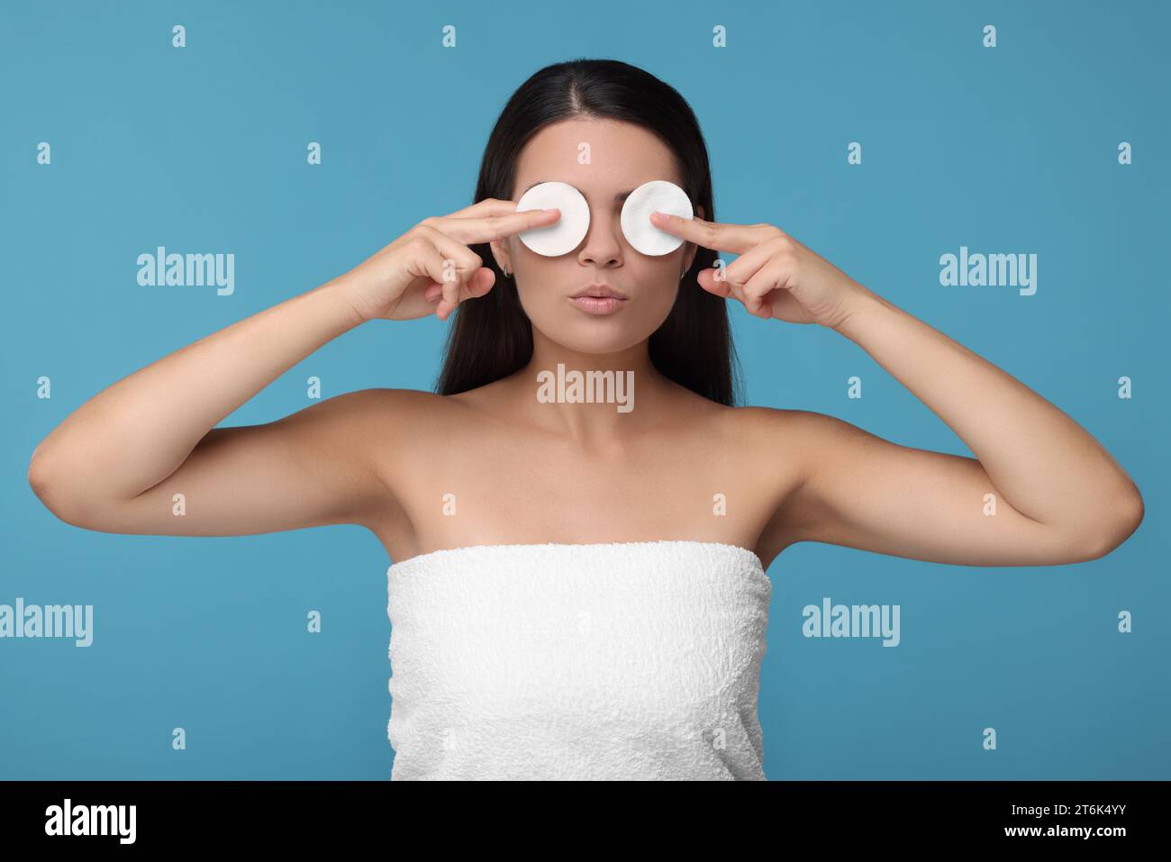 Young woman covering her eyes with cotton pads on light blue background ...