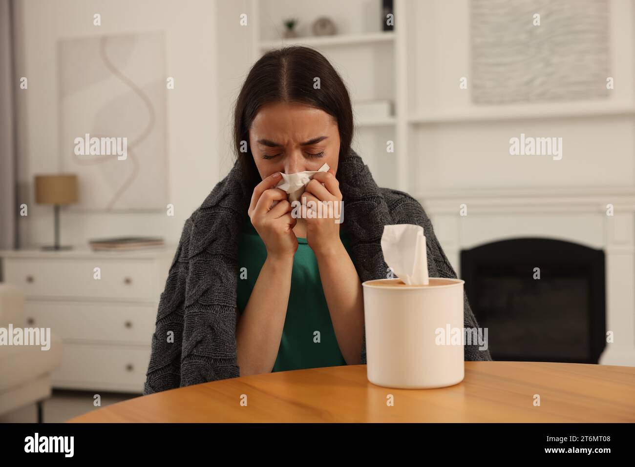 Sick woman wrapped in blanket with tissue blowing nose at wooden table ...