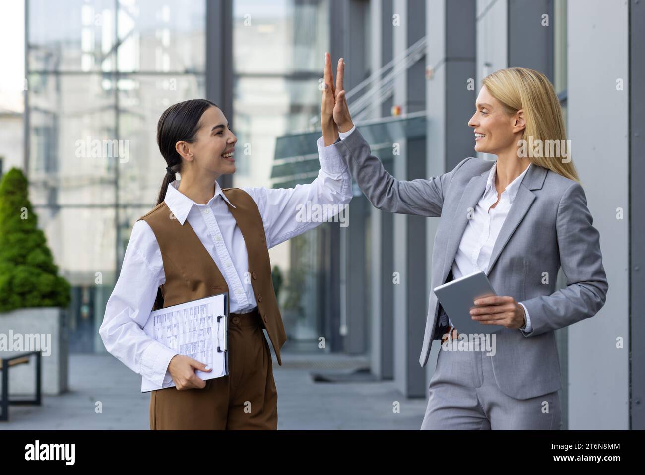 Two colleagues standing outside modern office, giving high fives to each other, happy after ...