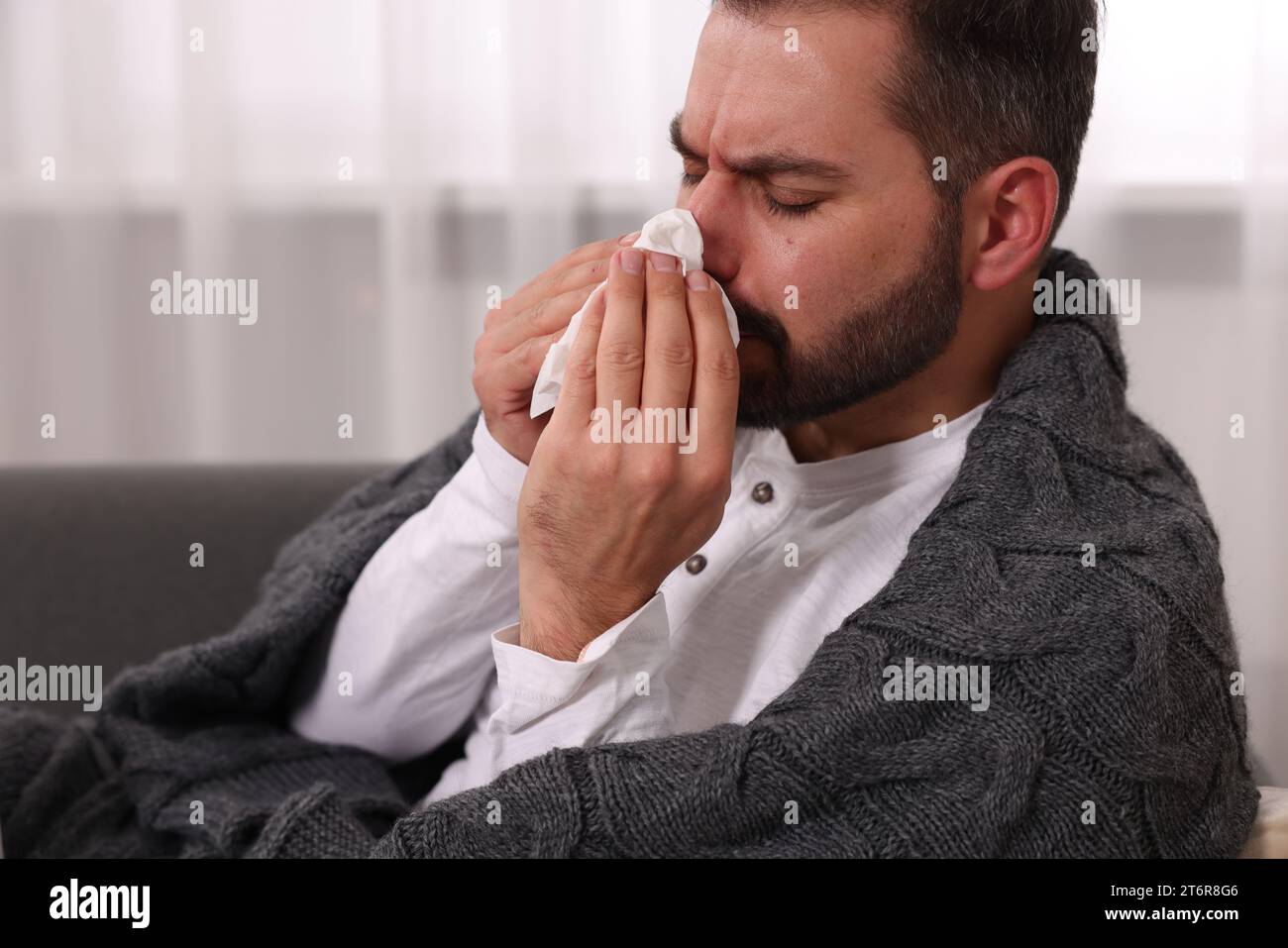 Sick man wrapped in blanket with tissue blowing nose on sofa indoors ...