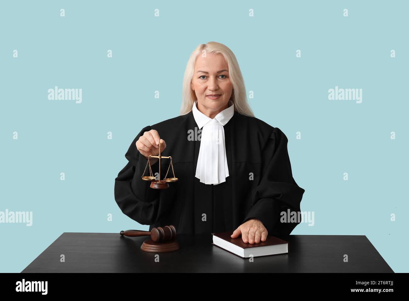 Mature female judge with scales of justice and book at table on blue ...