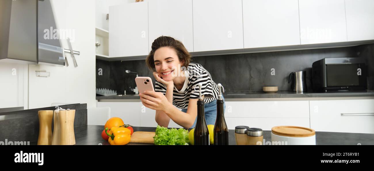 Portrait of smiling young woman cooking in the kitchen, watching videos ...