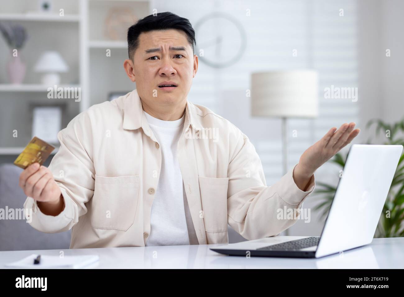 Confused asian man sitting in light office at table in front of laptop ...