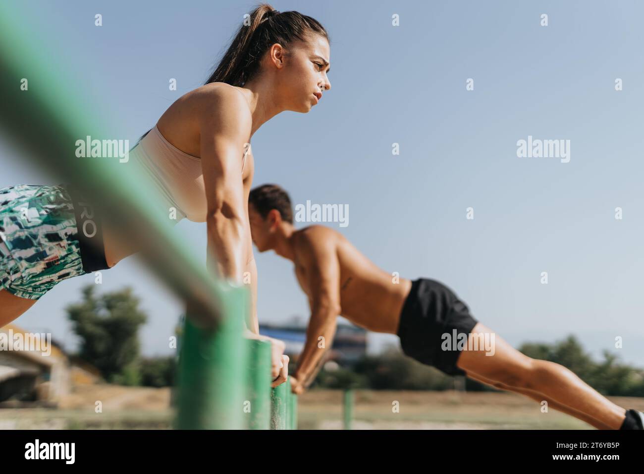 Fit couple exercising outdoors, doing pull-ups on metal bars in a sunny ...