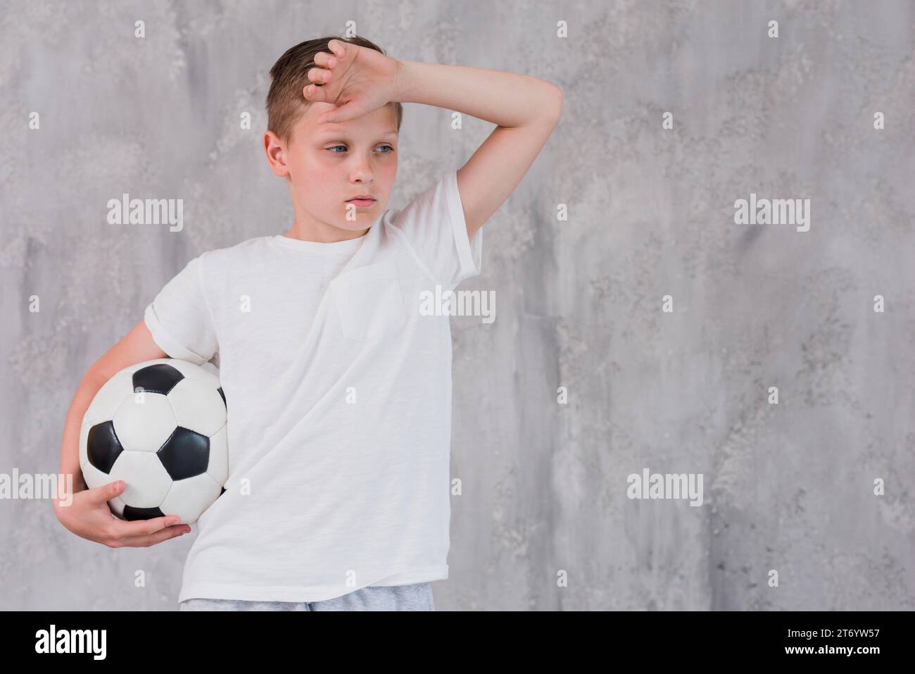 Portrait exhausted boy holding soccer ball hand against concrete wall ...