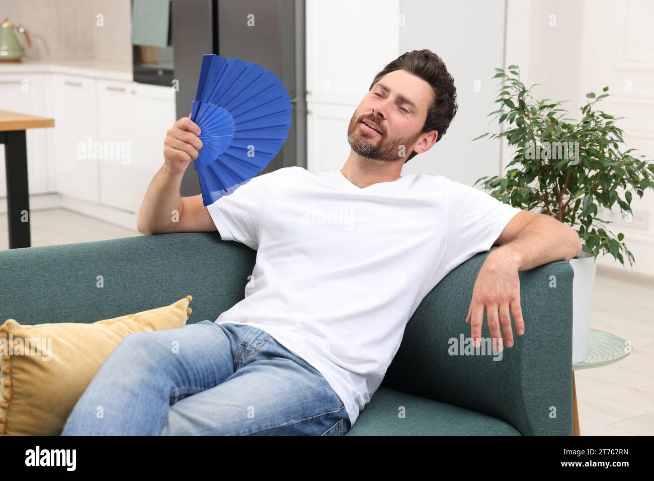 Bearded man waving blue hand fan to cool himself on sofa at home Stock ...