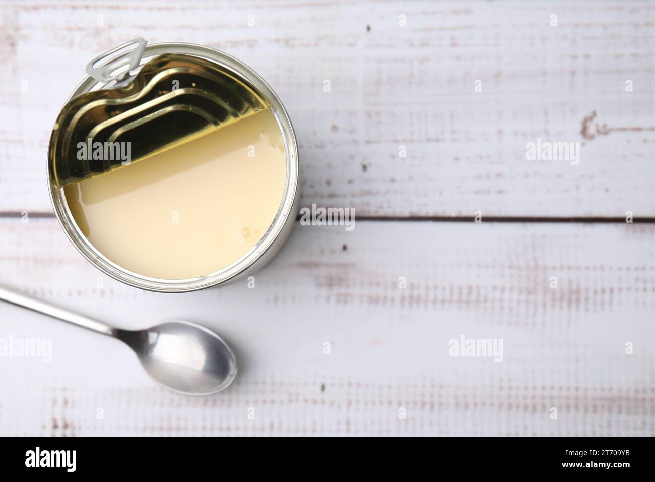 Tasty condensed milk in tin can and spoon on white wooden table, top ...