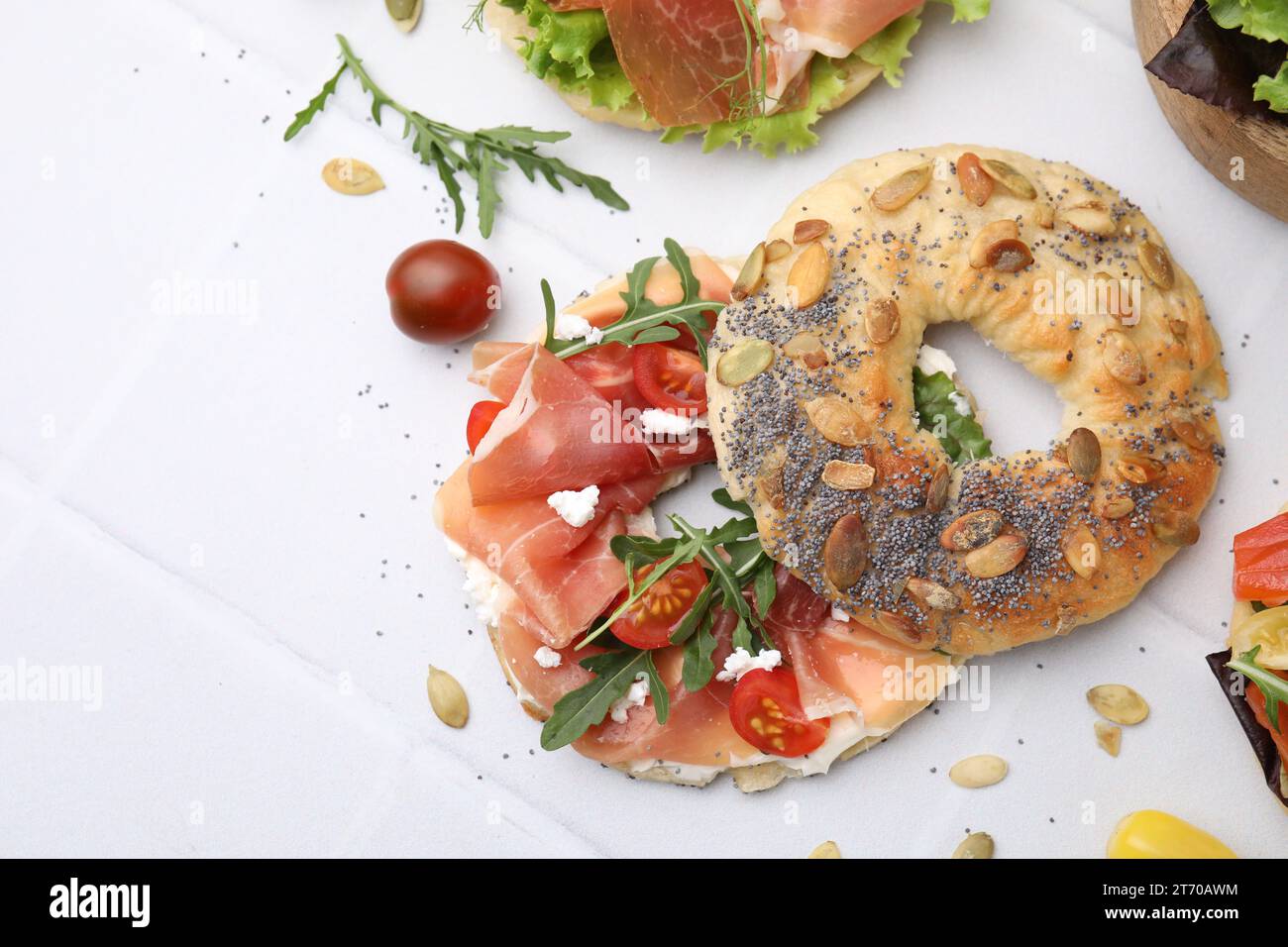 Tasty bagel with cured ham, cheese, tomatoes and arugula on white tiled ...