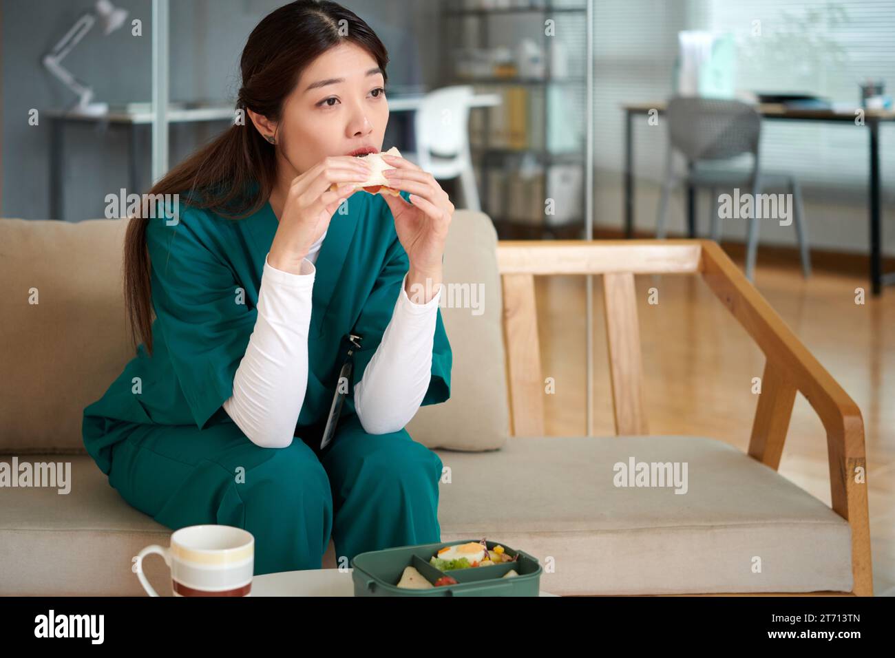 Tired young medical nurse eating sandwich when resting in hospital ...