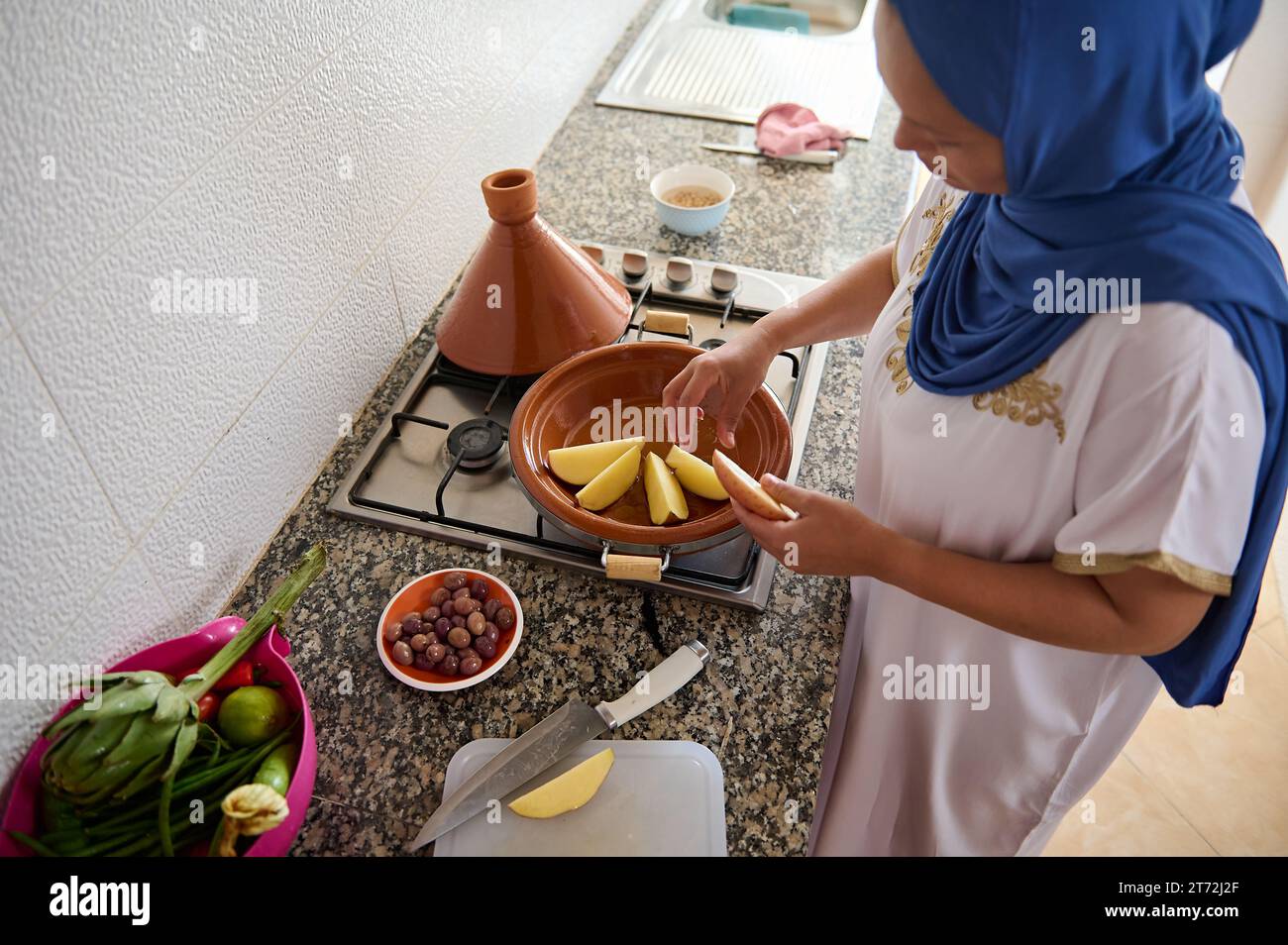 Authentic Arab Muslim woman in hijab and long dress, standing by stove on the kitchen counter ...