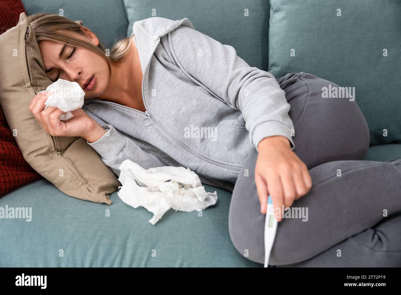 Young sick woman with cold and flu virus at home on sofa measuring body ...