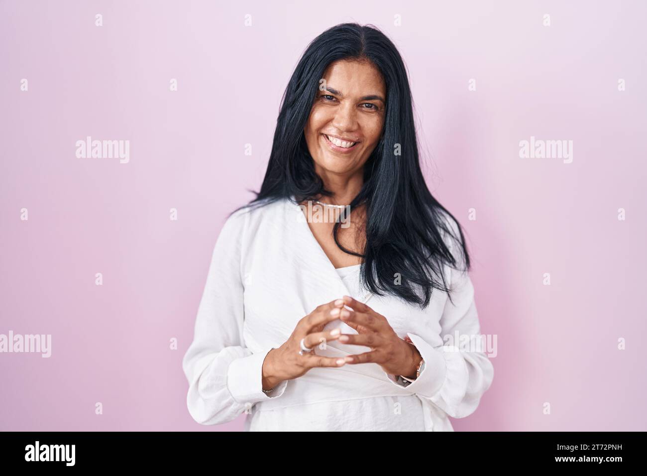 Mature hispanic woman standing over pink background hands together and ...