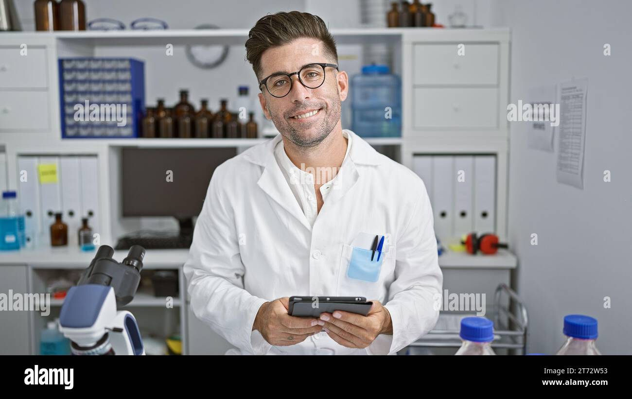 Handsome young hispanic man, confident smiling scientist, comfortably working with touchpad at ...