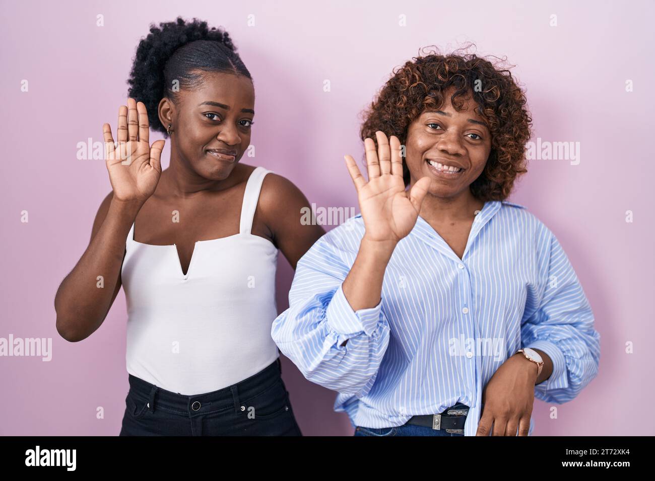 Two african women standing over pink background waiving saying hello ...
