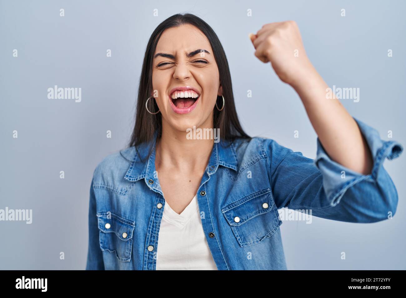 Hispanic woman standing over blue background angry and mad raising fist ...