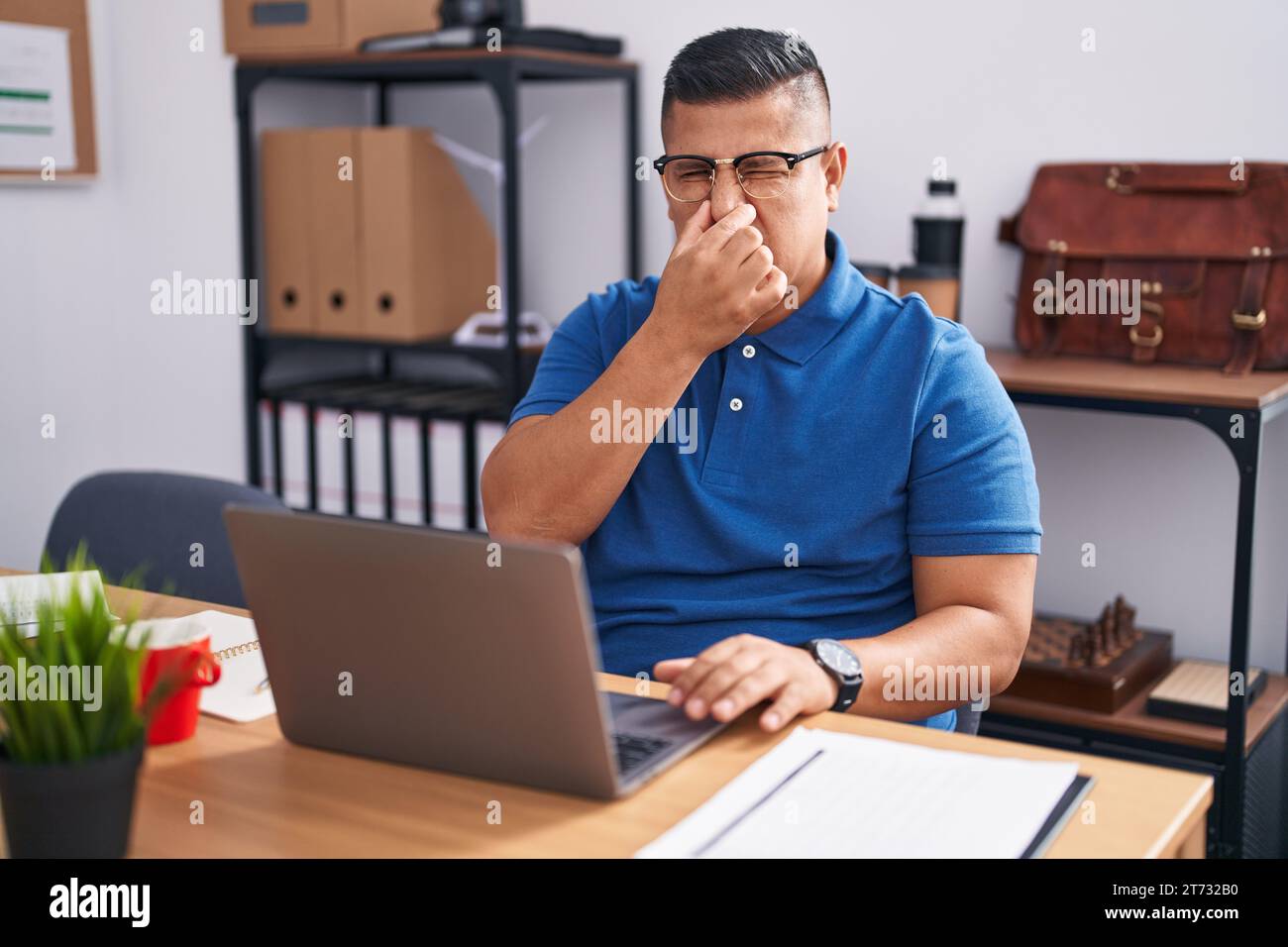 Young hispanic man working at the office with laptop smelling something ...