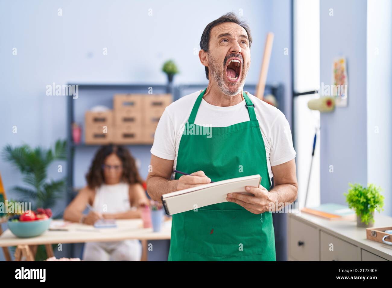 Middle age hispanic man painting on art notebook with pencils angry and ...