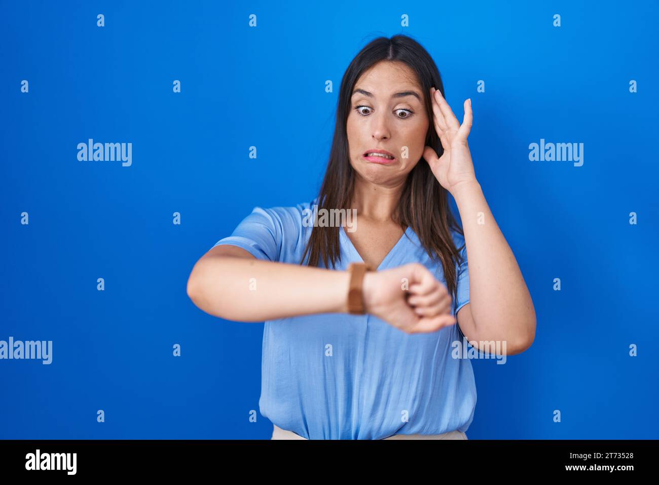 Young brunette woman standing over blue background looking at the watch ...