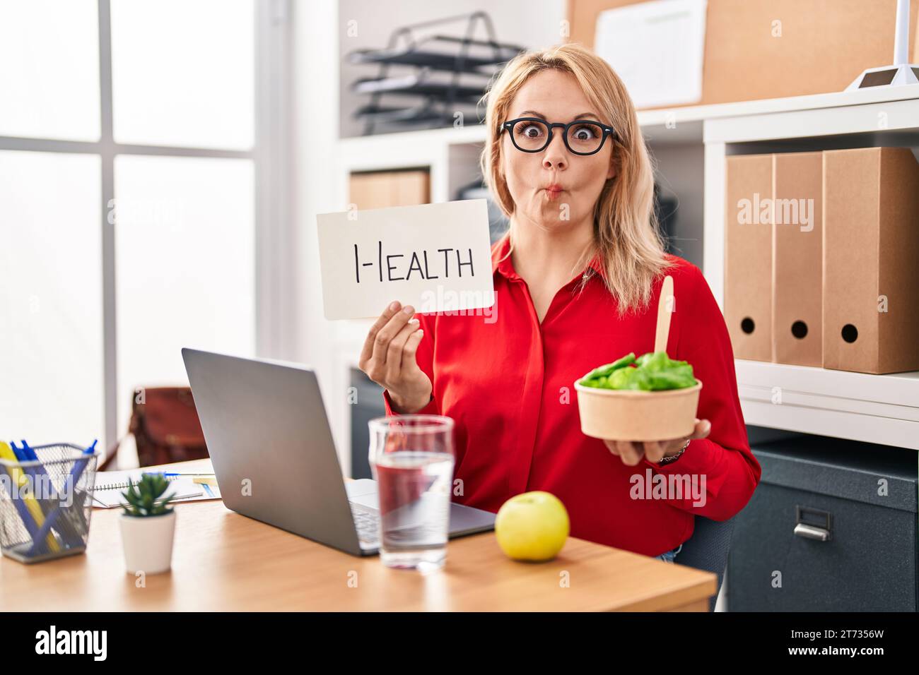 Blonde woman working at the office eating healthy food making fish face ...