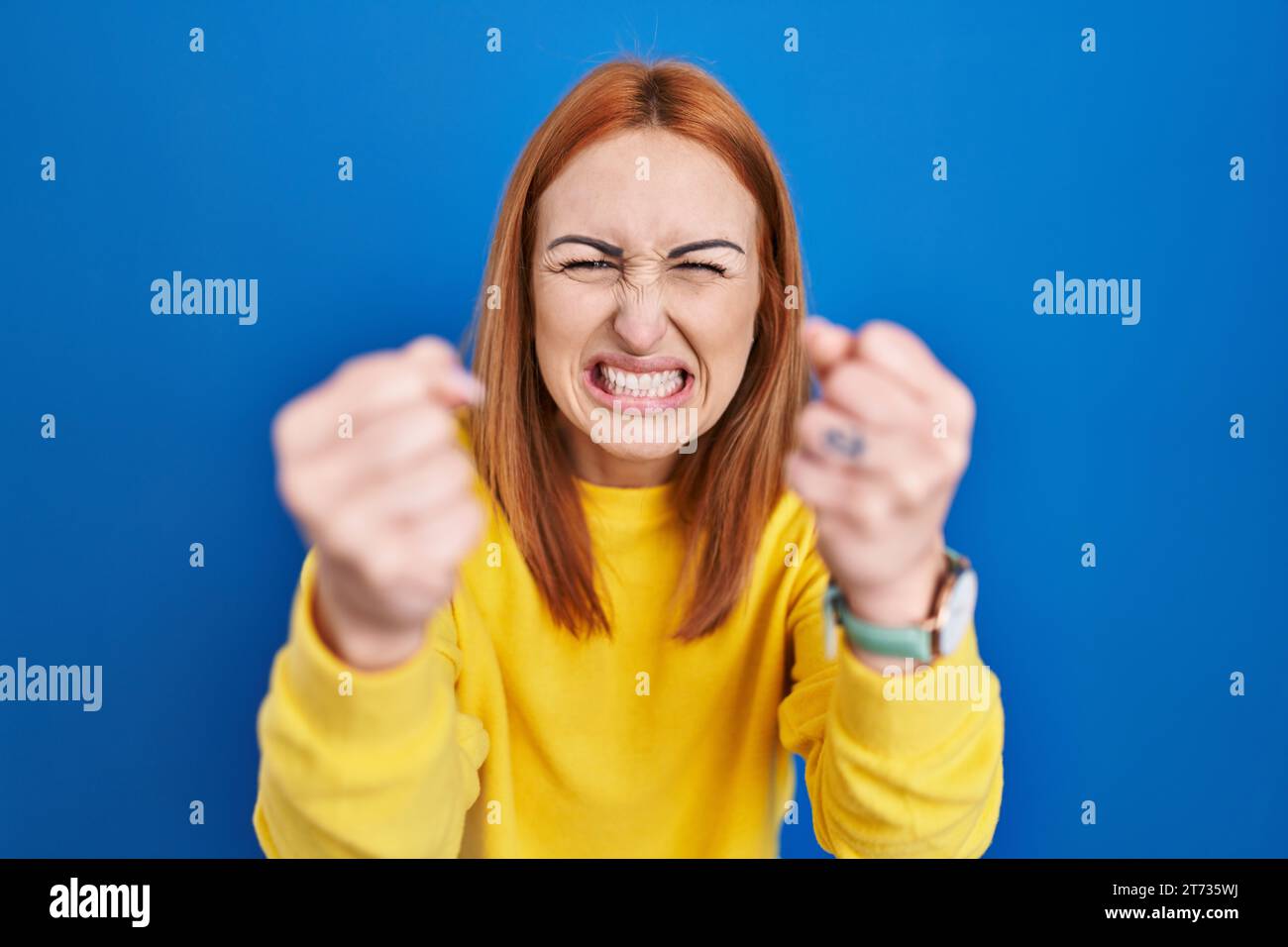 Young woman standing over blue background angry and mad raising fists ...